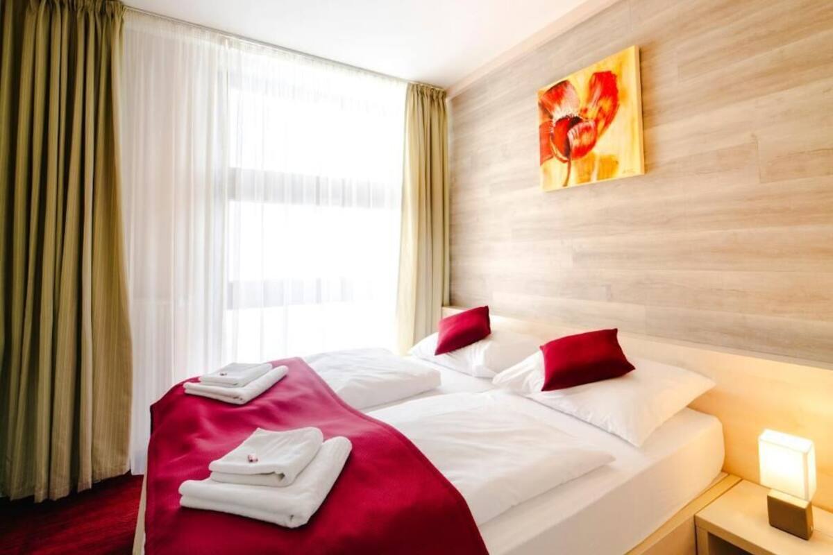 Quarto de hotel com uma cama grande, roupa de cama branca, almofadas vermelhas e um cobertor vermelho. Há uma pintura na parede e uma mesinha de cabeceira com uma luminária.