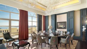Quarto luxuoso com uma grande mesa de jantar posta para seis pessoas, rodeada por cadeiras elegantes. Um lustre pendurado no teto e uma ampla janela oferecem vista para o mar.