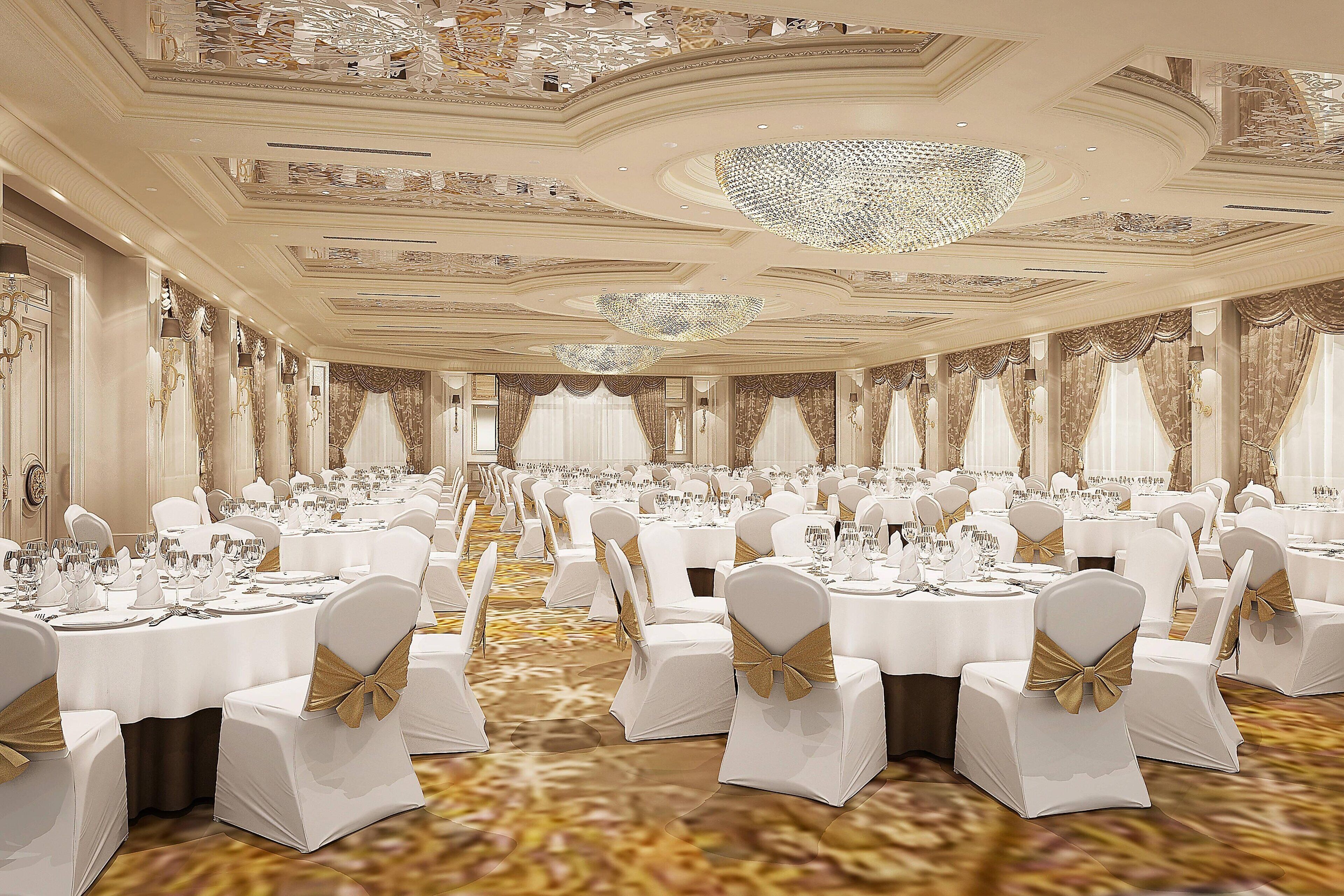 Uma sala de banquetes com mesas redondas, cadeiras brancas com fitas douradas e decorações ornamentais no teto.