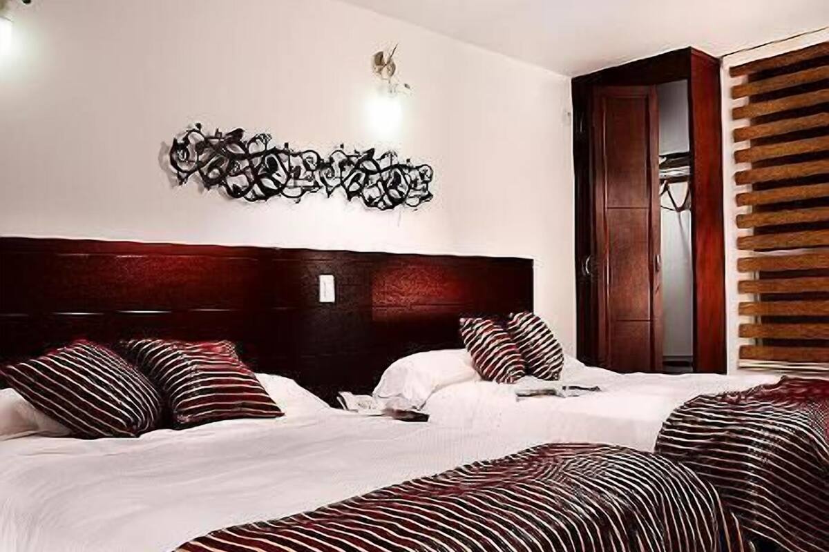 Quarto de hotel com cabeceira de madeira, duas camas com roupa de cama listrada, uma obra de arte abstrata na parede e um guarda-roupa de madeira.