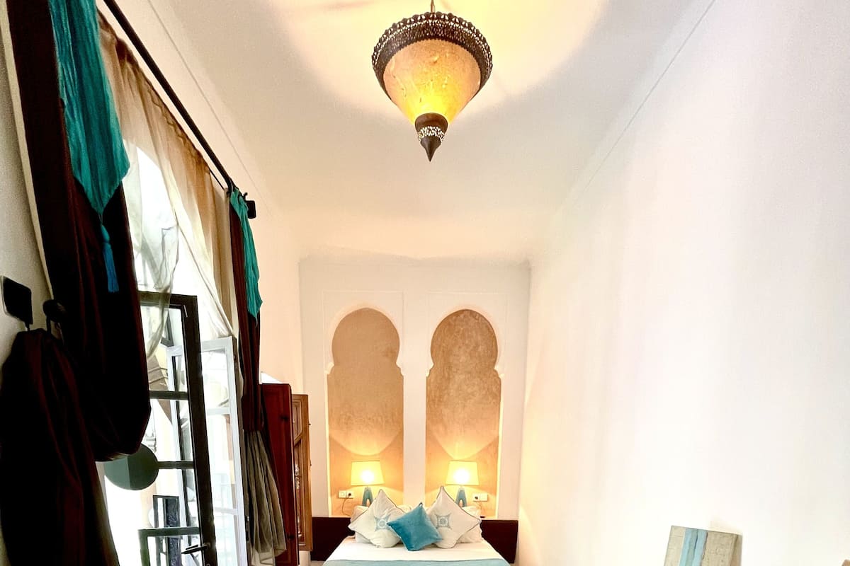 Quarto de hotel com cama, escrivaninha, cadeira, mesinha, luminária pendente e vista para o exterior.