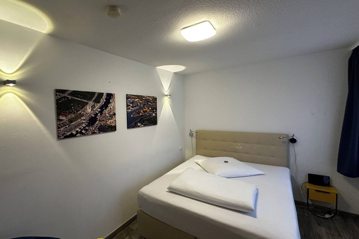 Quarto de hotel com uma cama, duas luminárias na parede e duas fotografias emolduradas.