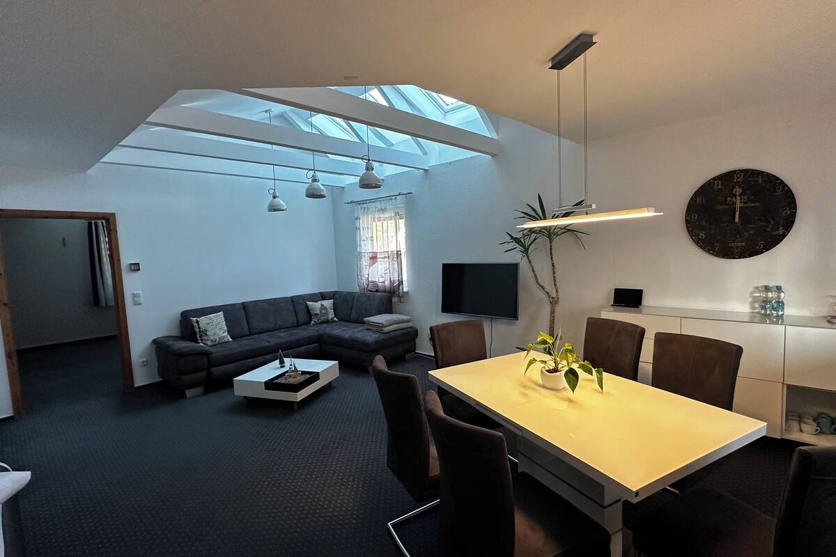 Uma sala de estar moderna com teto alto, claraboia e área de jantar com mesa e cadeiras.