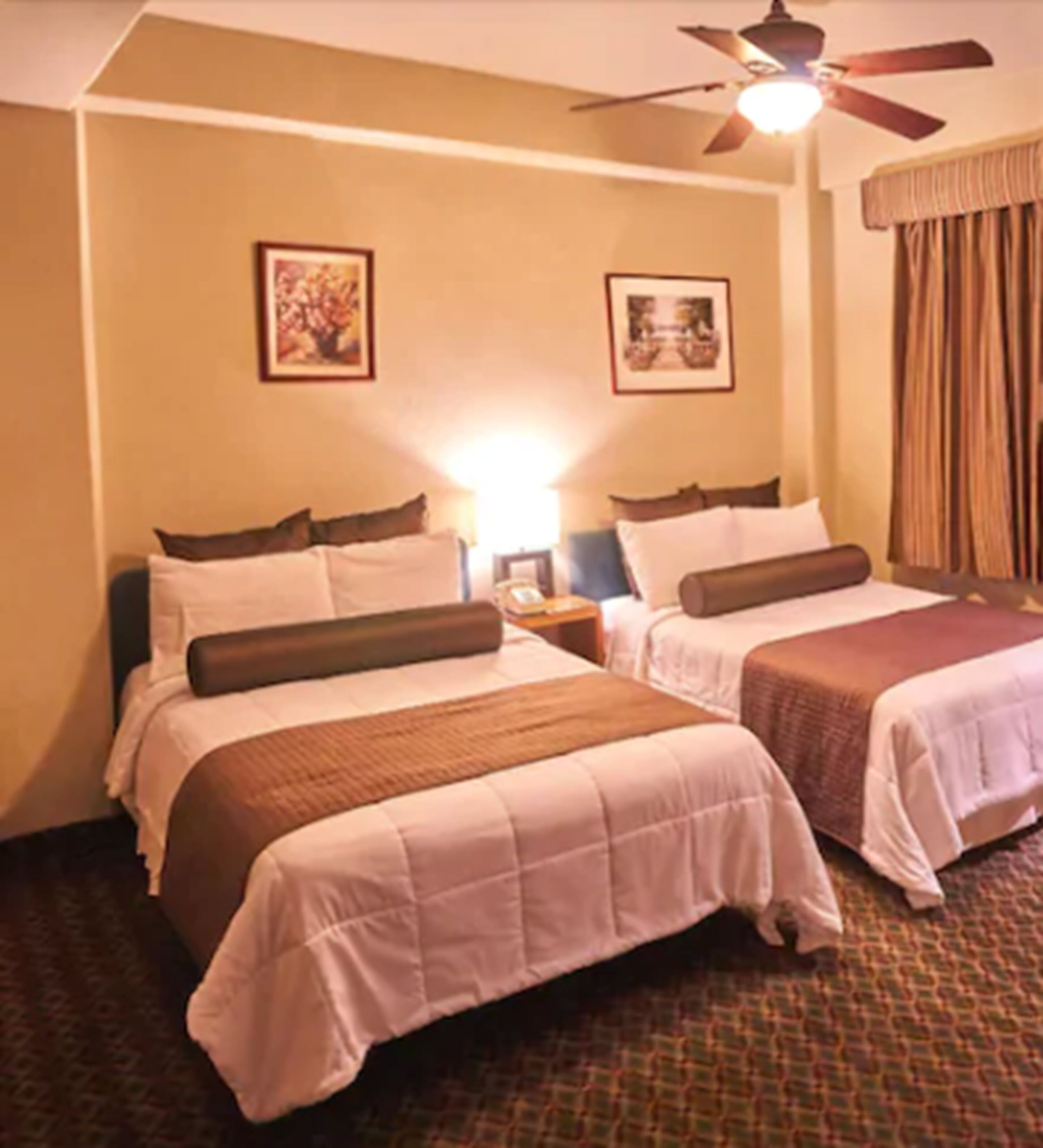 Quarto de hotel com duas camas, um ventilador de teto e duas fotos emolduradas na parede.