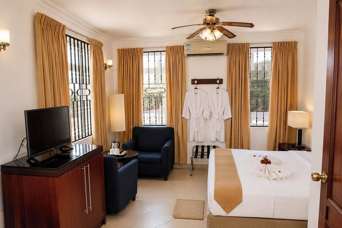 Quarto de hotel com cama, escrivaninha com televisão, cadeira, ventoinha no teto e janela com cortinas.