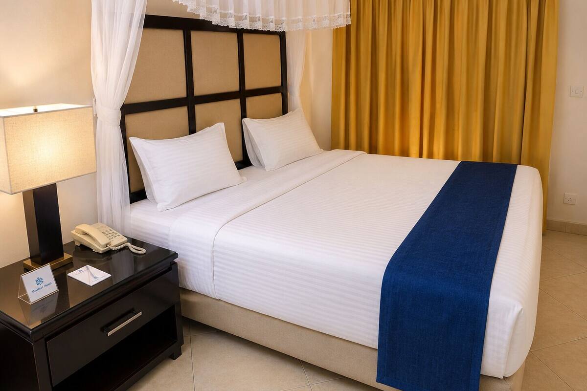 Quarto de hotel com cama, mesa de cabeceira, abajur, telefone e placa com o nome do hotel.