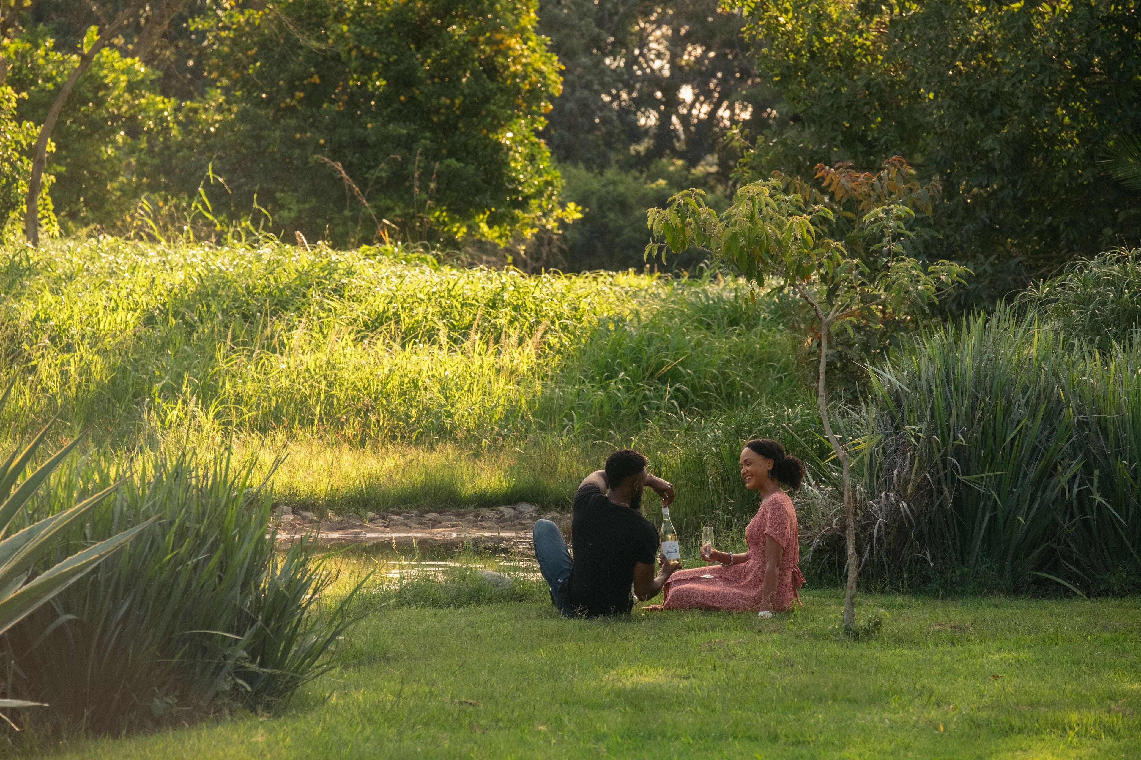 Duas pessoas sentadas na grama em ambiente natural, com árvores e vegetação ao fundo.