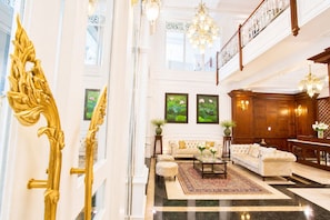 Um hall imponente com teto alto, lustres, uma escadaria monumental e uma área de estar com uma mesa de centro e sofás.