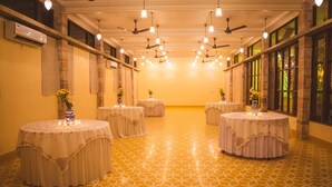 Um salão amplo com várias mesas redondas cobertas por toalhas brancas, cada uma decorada com velas e arranjos florais. O espaço possui um piso amarelado estampado e grandes janelas com molduras escuras.