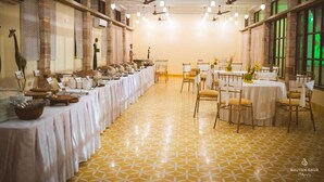 Salão de banquetes com longas mesas postas para refeições, ventiladores no teto e grandes janelas.