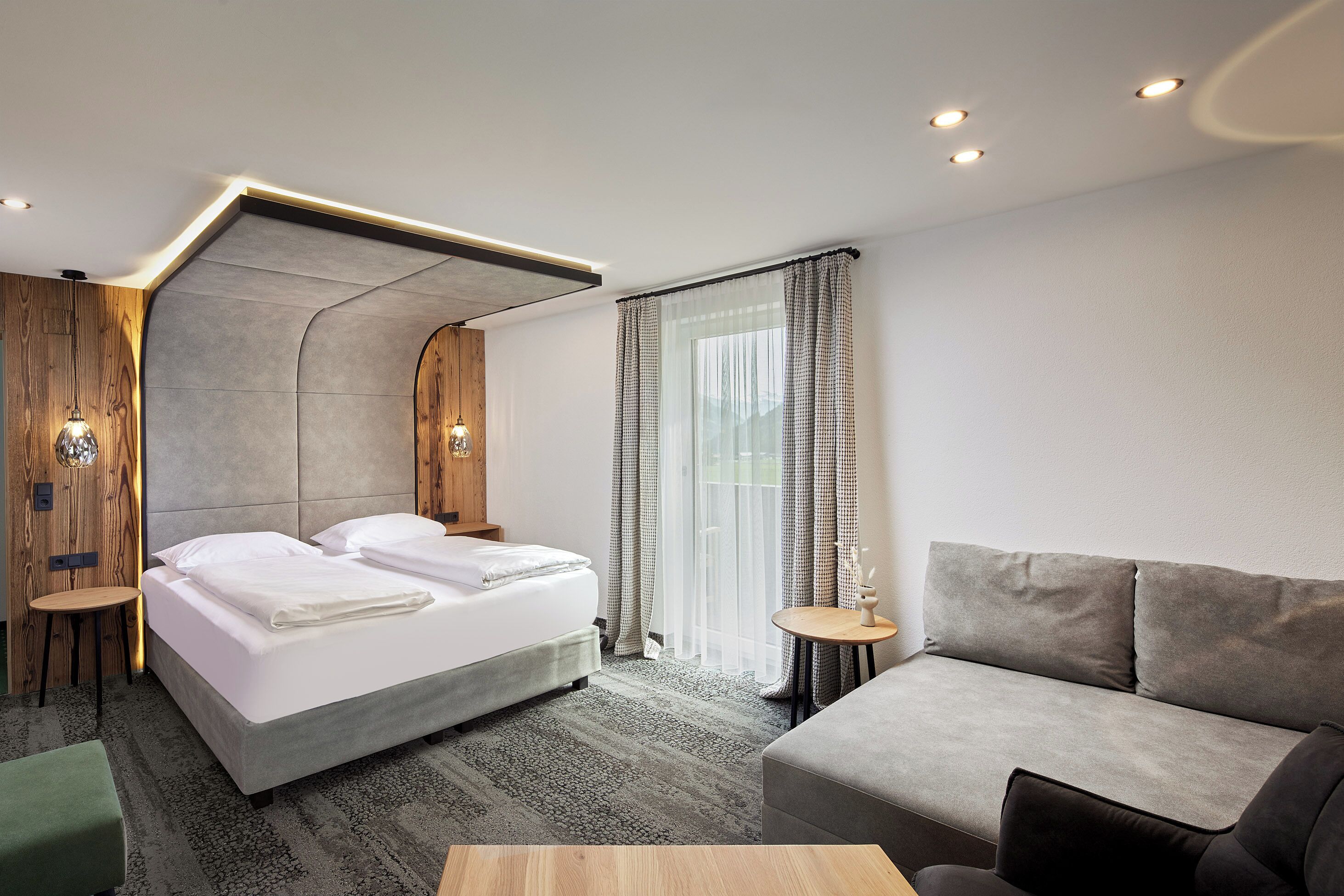 Quarto de hotel moderno com uma cama grande, um sofá, uma mesinha e uma janela com cortinas.
