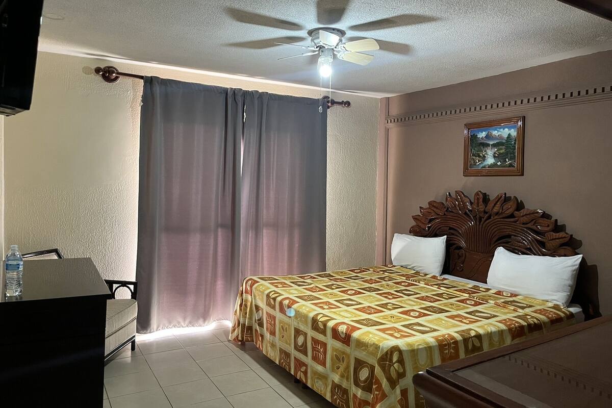 Quarto de hotel com cama, escrivaninha, ventoinha no teto, janela com cortinas e um quadro na parede.