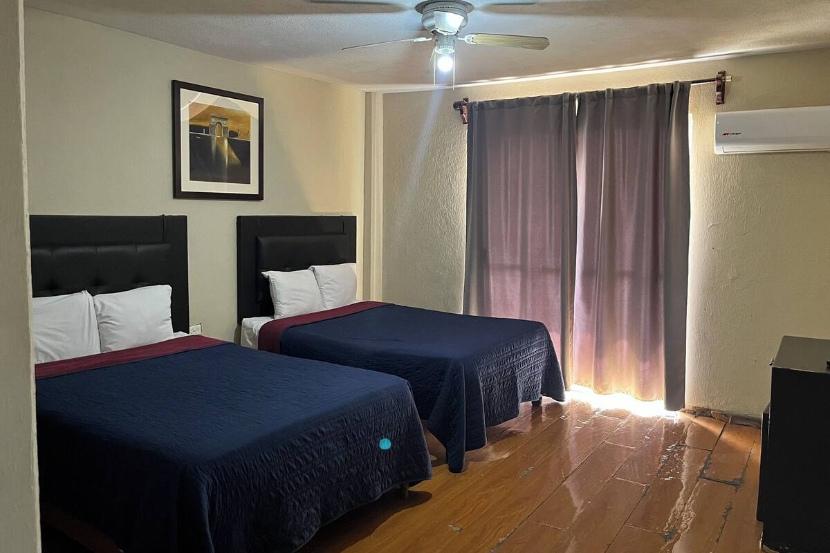 Quarto de hotel com duas camas, um ventilador de teto, uma moldura com imagem na parede e uma janela com cortinas.