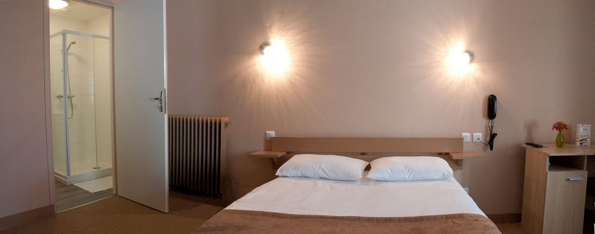 Quarto de hotel com cama, luminárias de cabeceira, telefone, radiador e área de chuveiro.