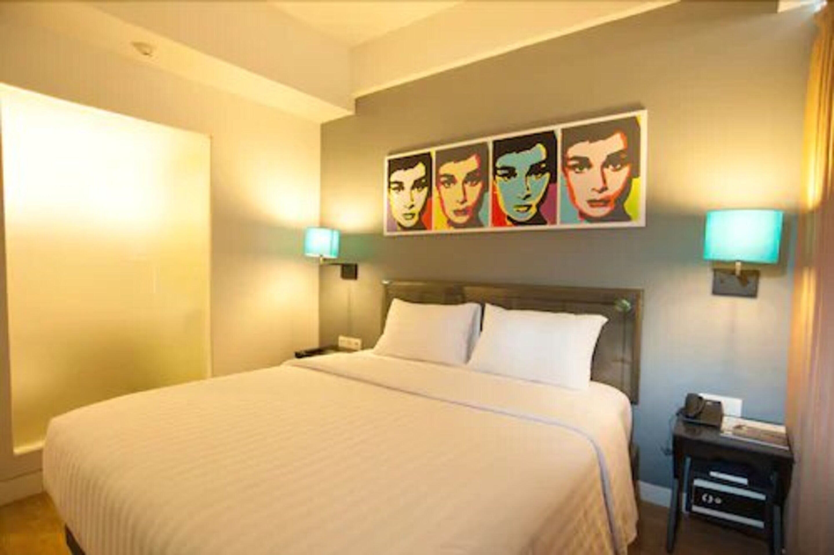Quarto de hotel com uma cama grande, cabeceira, abajures e uma parede com três obras de arte emolduradas.