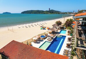 Um resort à beira-mar com piscina, cadeiras de praia, guarda-sóis e vista para o mar e colinas ao longe.