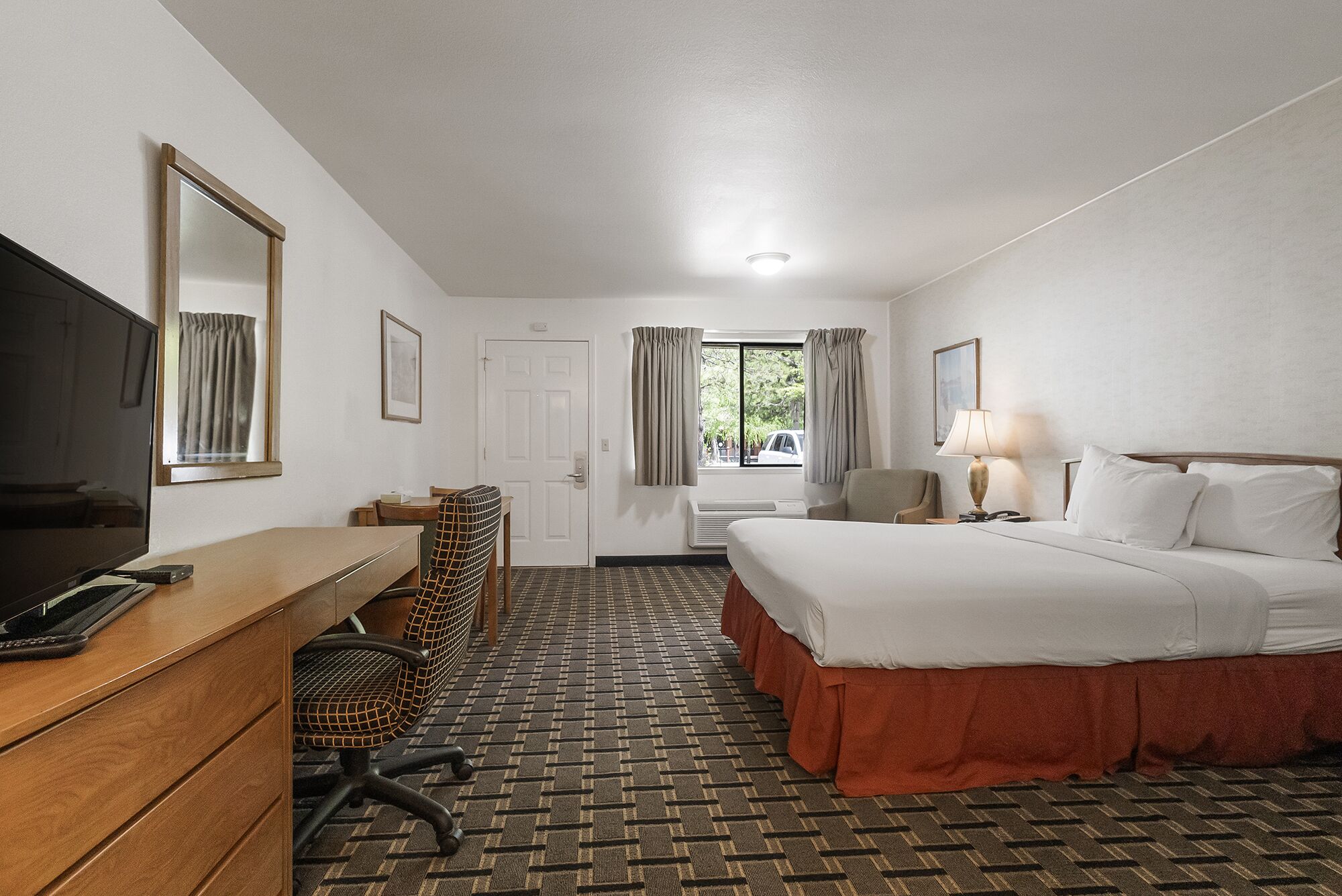 Quarto de hotel com cama, escrivaninha, cadeira, televisão e janela com cortinas.