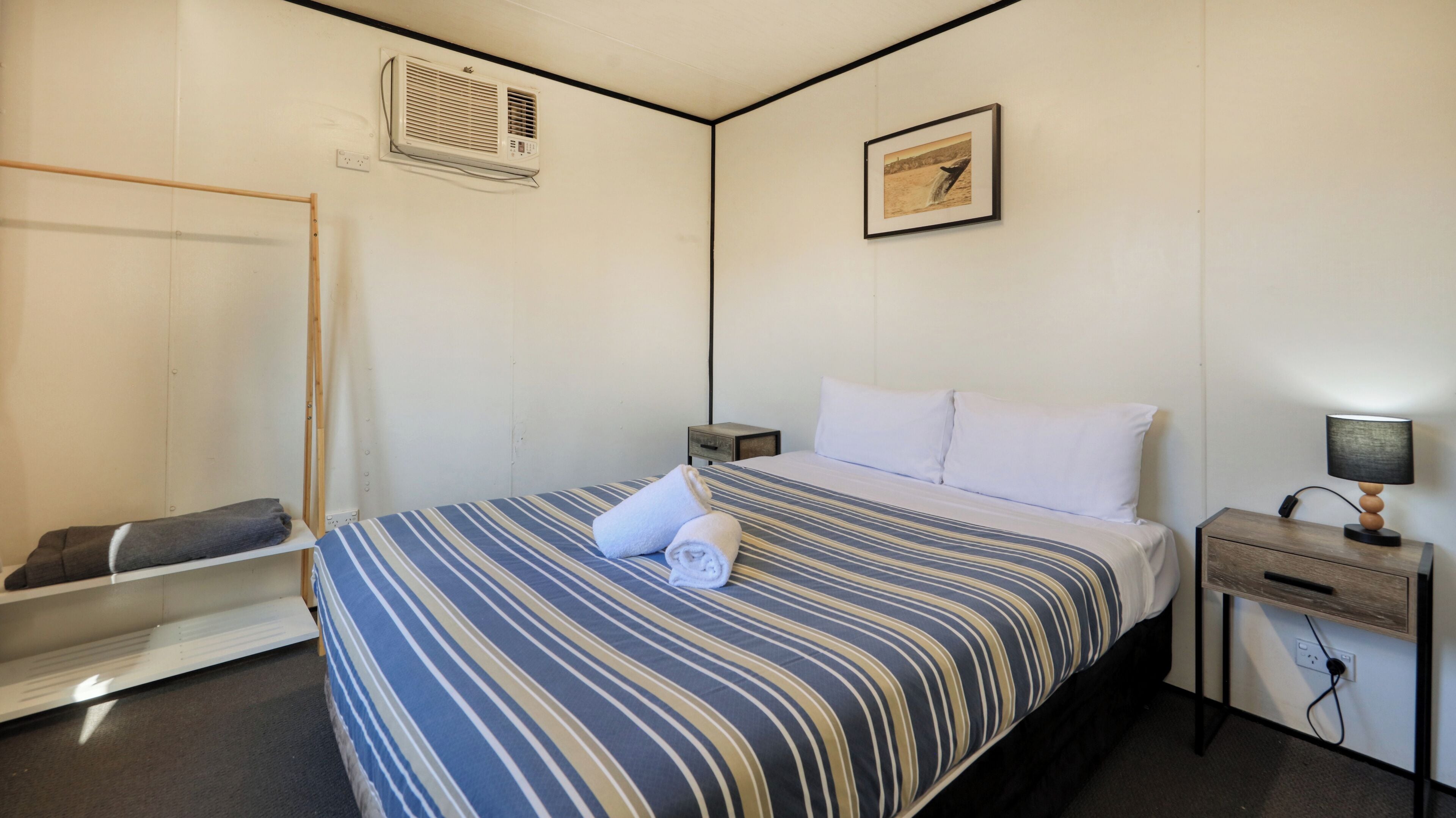 Quarto de hotel com cama, mesa de cabeceira, luminária, quadro na parede e guarda-roupa.