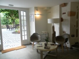 Uma sala de estar moderna com uma mesa redonda, duas cadeiras, um abajur e uma porta que dá para um pátio externo com piso de tijolos e mobiliário de exterior.