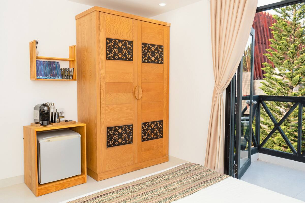 Quarto com guarda-roupa de madeira, um pequeno frigorífico, uma estante com livros e uma máquina de café, e vista para uma varanda com grades e vegetação.