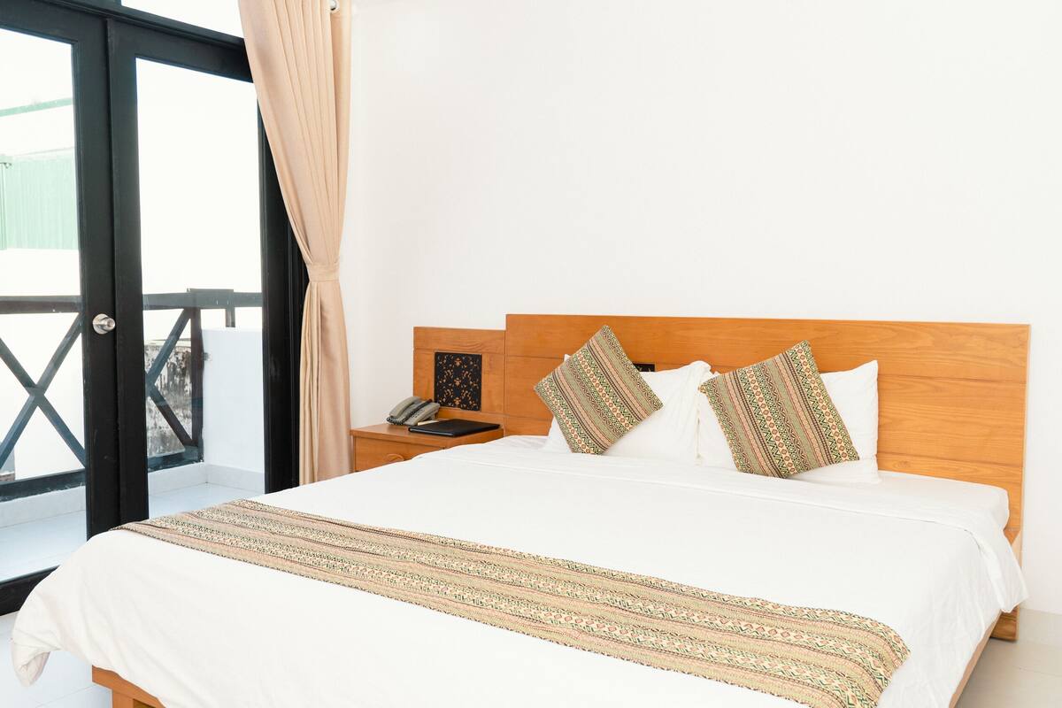 Quarto de hotel com uma cama grande, cabeceira de madeira, duas almofadas estampadas, mesa de cabeceira com telefone e um caderno, e vista através de uma porta de vidro deslizante.