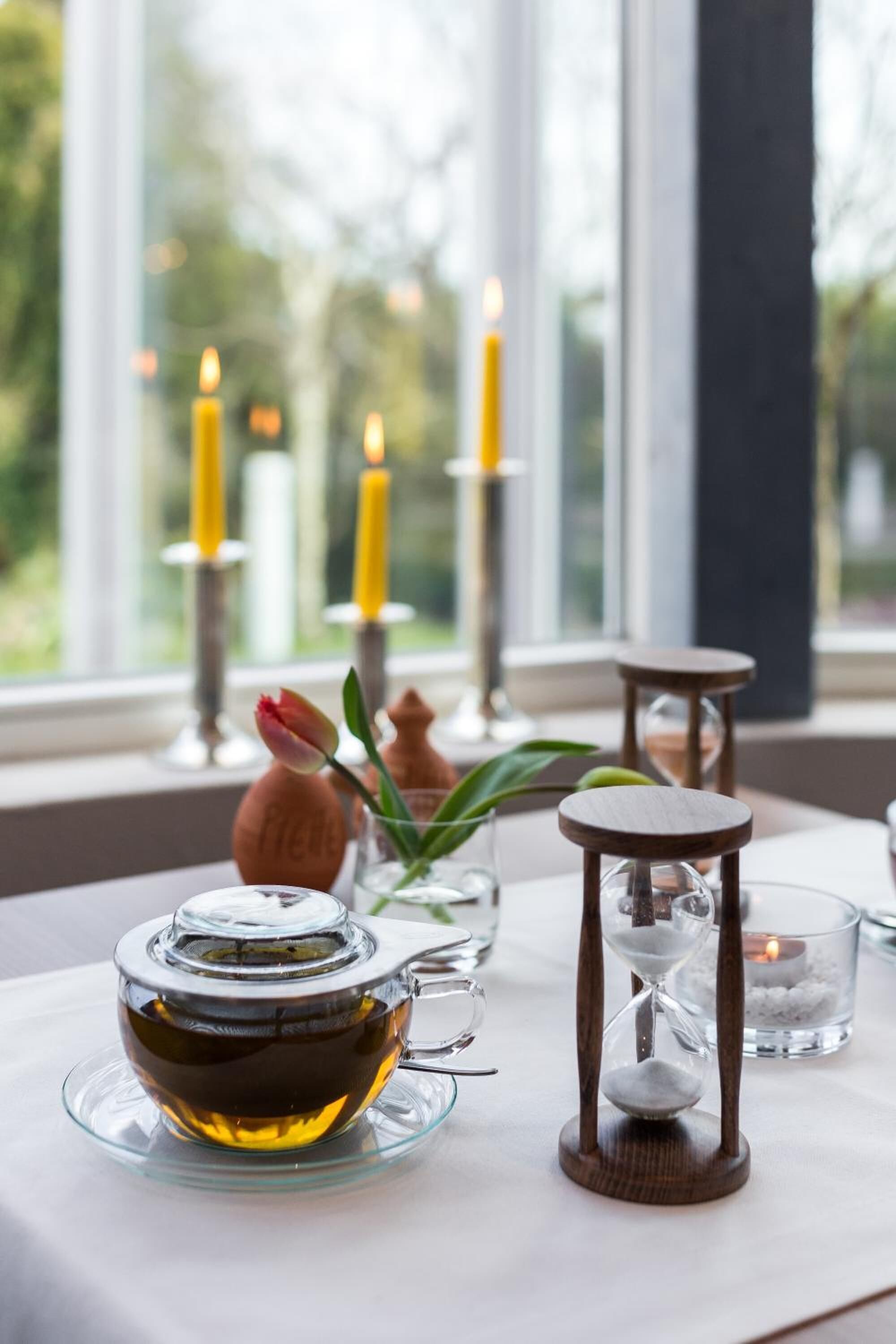 Uma mesa com um bule, um ampulheta, velas e um vaso de flores.