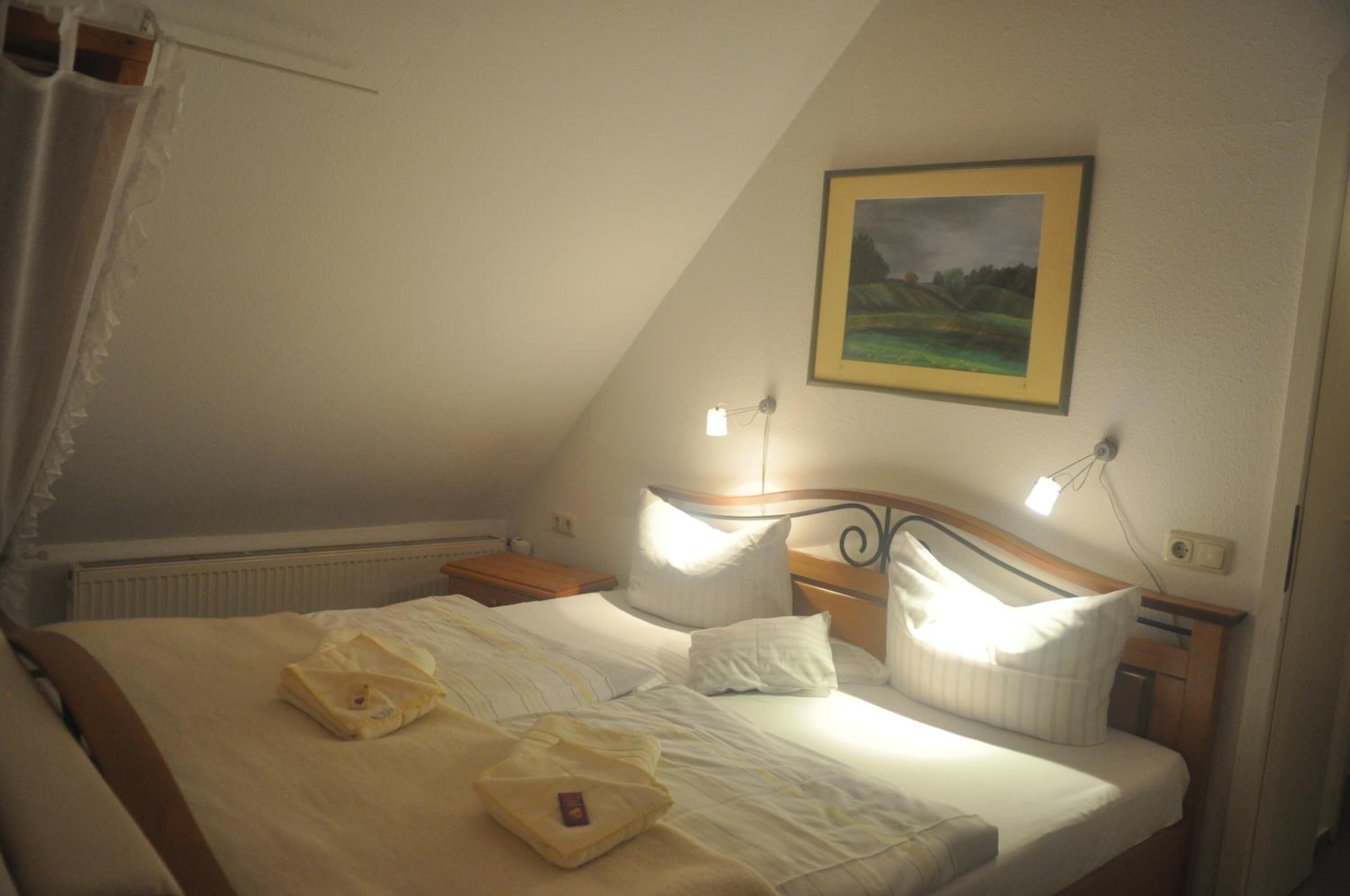Cama bem arrumada com lençóis brancos, duas luminárias de cabeceira, um quadro com uma paisagem e uma mesinha de cabeceira de madeira.
