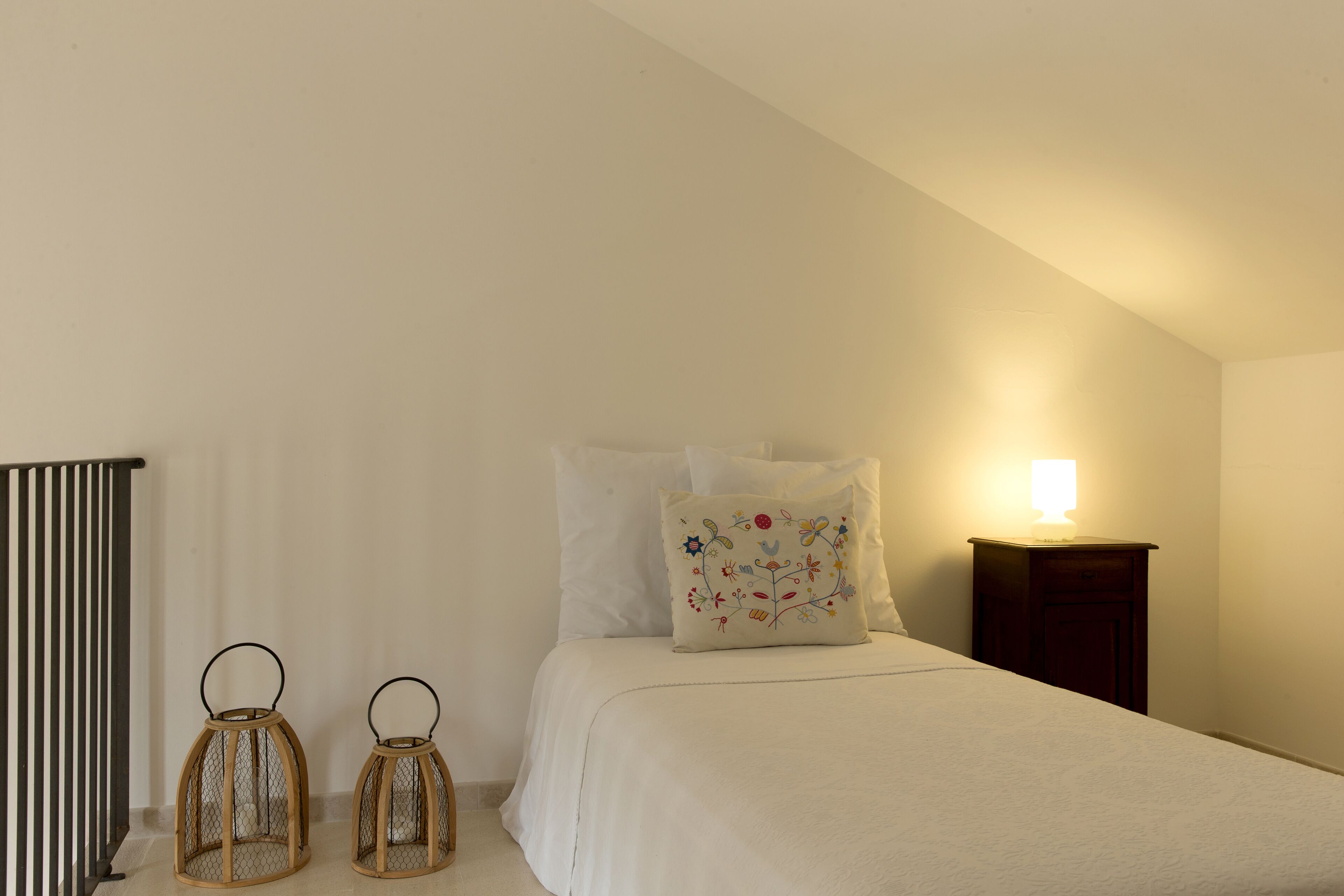 Quarto com cama, roupa de cama branca, mesa de cabeceira com abajur e duas lanternas decorativas.