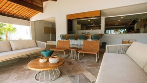 Uma área de estar moderna com sofá, cadeiras de vime, uma mesa redonda de madeira e um bar com banquetas.