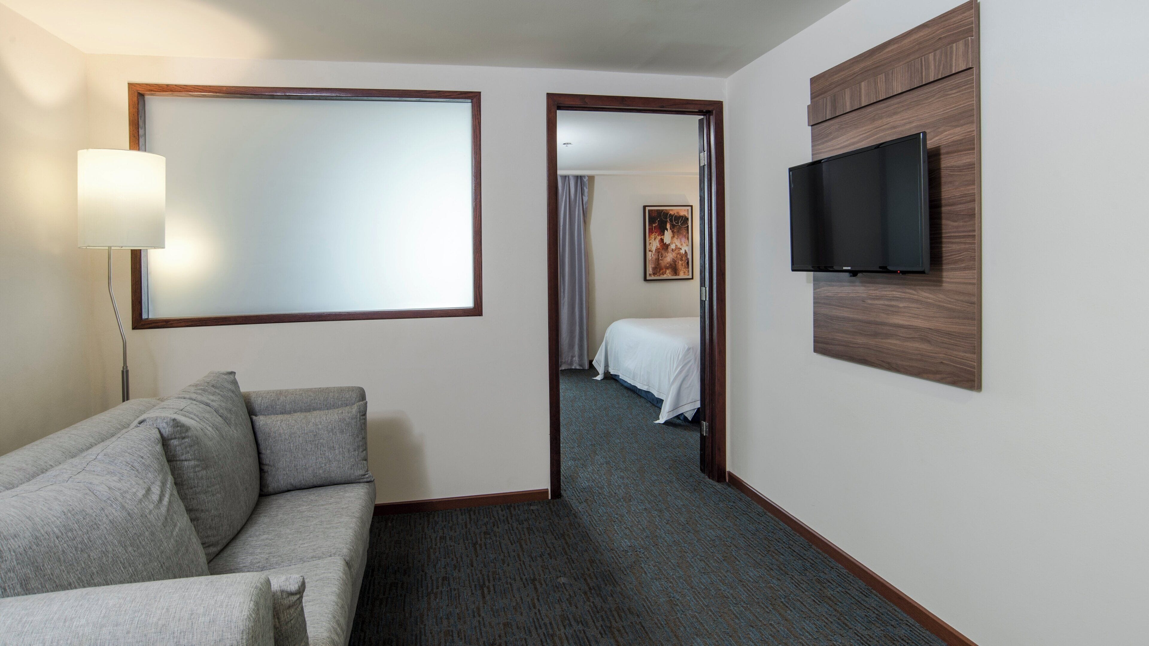 Quarto de hotel com sofá, TV de tela plana, espelho grande e porta que dá para um quarto com cama e um quadro.