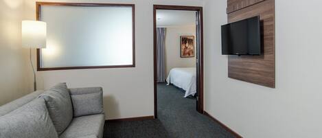 Quarto de hotel com sofá, TV de tela plana, espelho grande e porta que dá para um quarto com cama e um quadro.