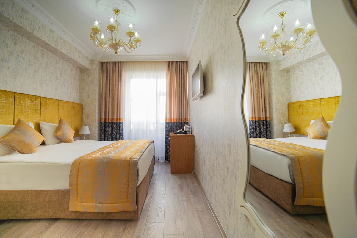 Quarto de hotel com duas camas, cabeceira de madeira, uma mesinha de cabeceira pequena, televisão fixada na parede e um espelho refletindo parte do quarto.