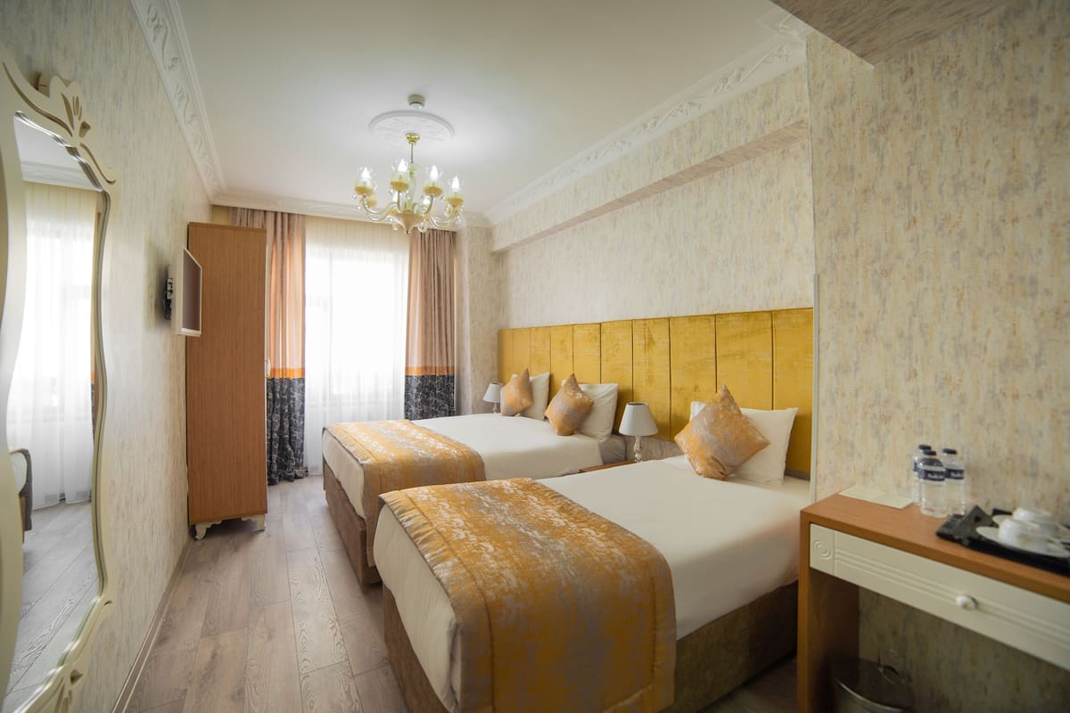Quarto de hotel com duas camas, cabeceira de madeira, escrivaninha e lustre.