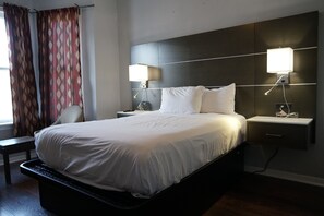 Uma cama bem arrumada com lençóis brancos, uma mesa de cabeceira com abajur e um criado-mudo também com abajur.