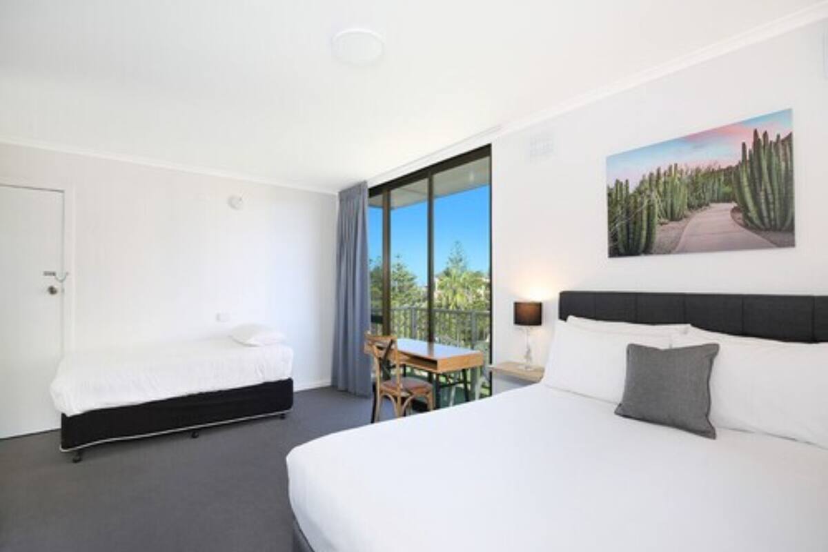 Quarto de hotel com uma cama grande, uma cama menor, uma escrivaninha e uma cadeira. Há uma janela com vista para árvores e um quadro de cactos na parede.