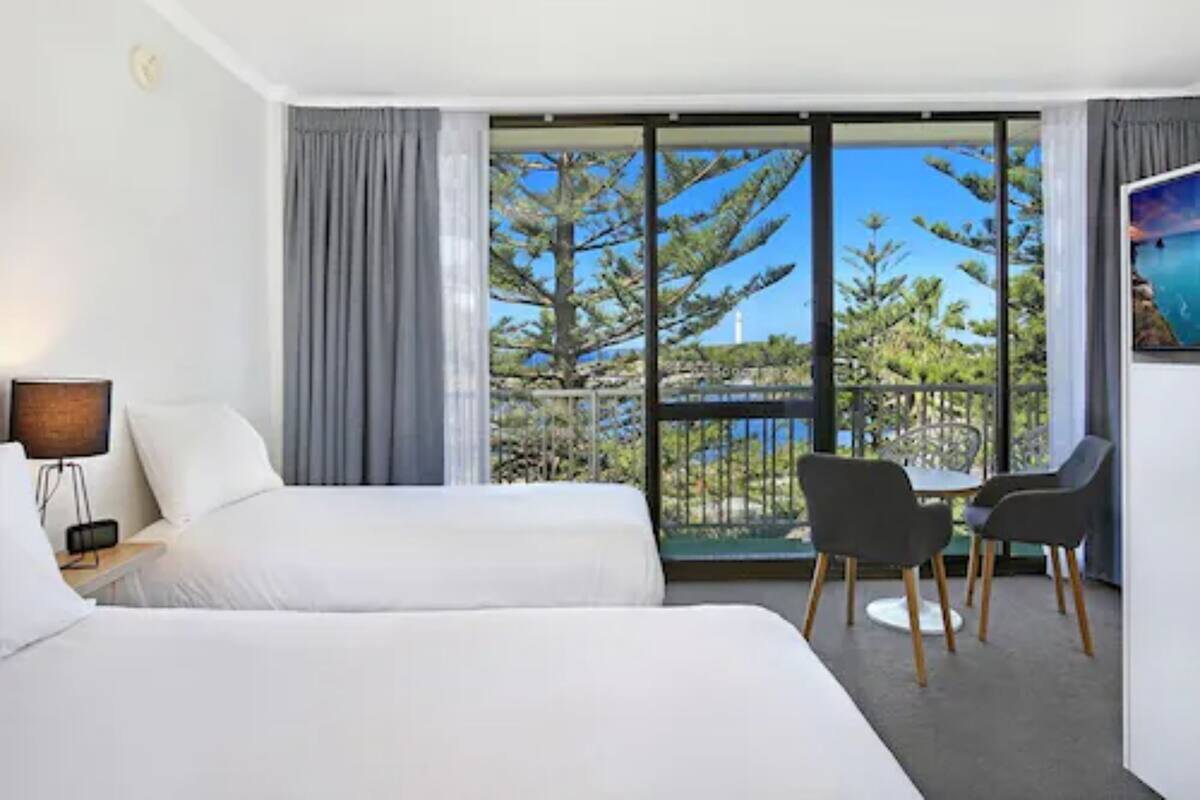 Quarto de hotel com duas camas, varanda com mesa e cadeiras, e vista para árvores e um corpo d'água.