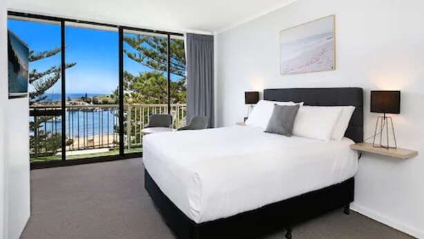 Quarto de hotel moderno com uma cama grande, vista para o mar e uma varanda com guarda-corpo.