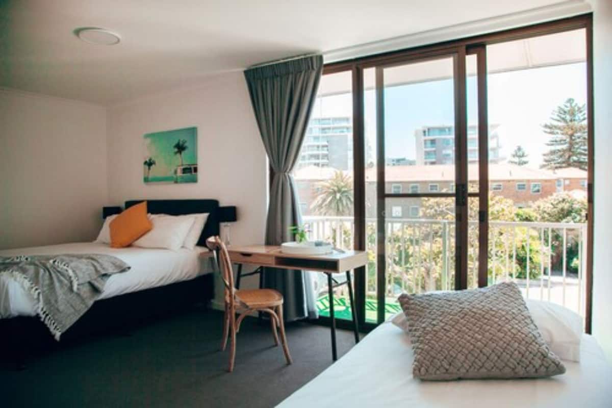 Quarto de hotel com cama, escrivaninha, cadeira e uma janela grande com vista para edifícios e vegetação.