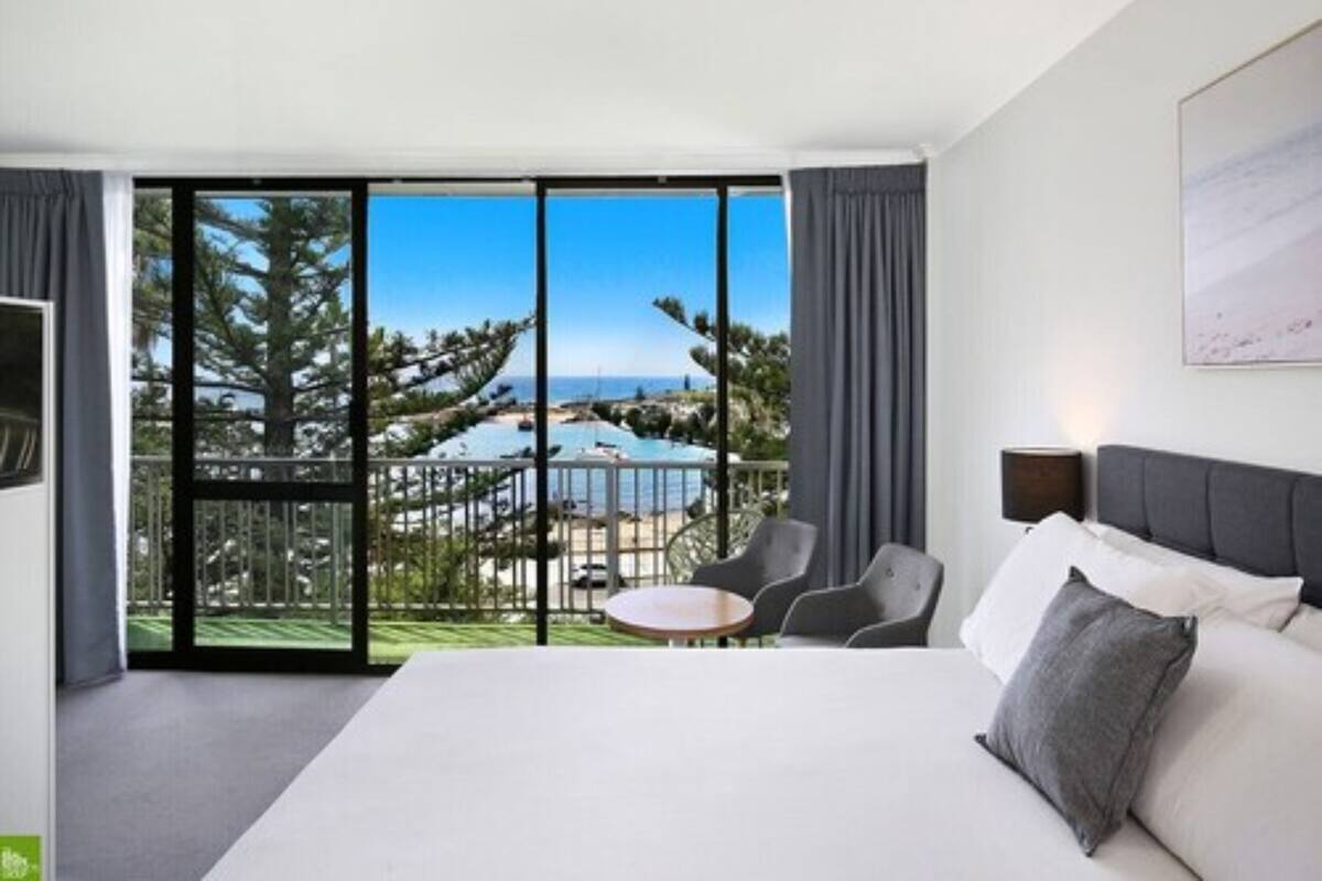 Quarto de hotel moderno com uma cama grande, uma mesa pequena e vista para uma piscina e árvores.
