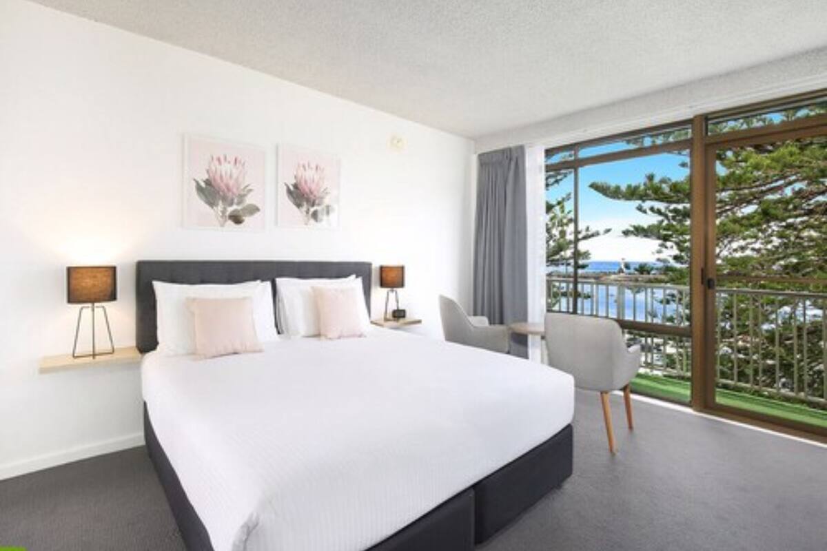 Quarto de hotel moderno com uma cama grande, uma escrivaninha e uma cadeira. Há duas luminárias fixadas na parede e uma janela ampla com vista para o exterior.