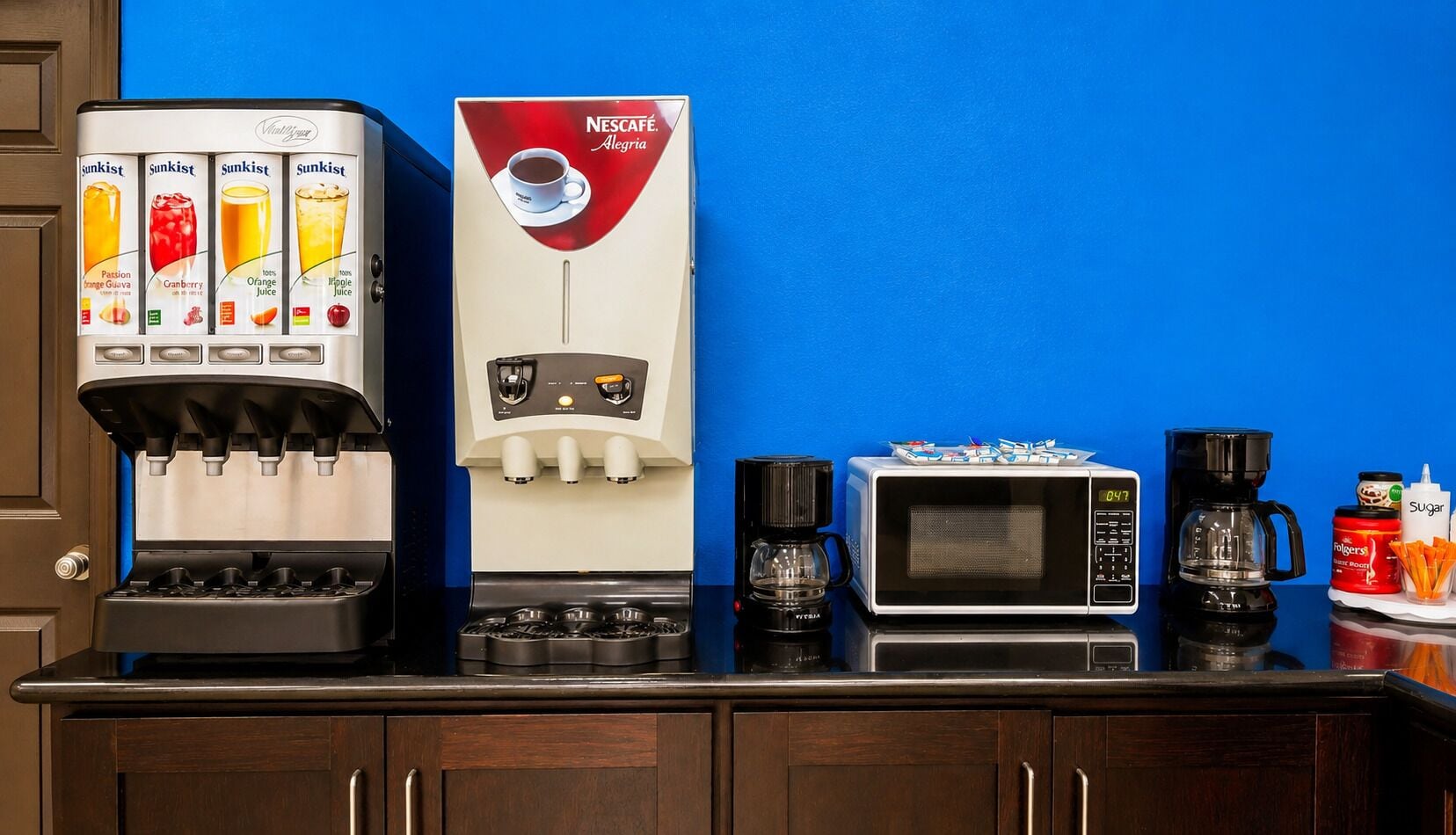 Uma bancada de cozinha com um dosador de suco Sunkist, uma máquina de café Nescafé, um micro-ondas e uma cafeteira.