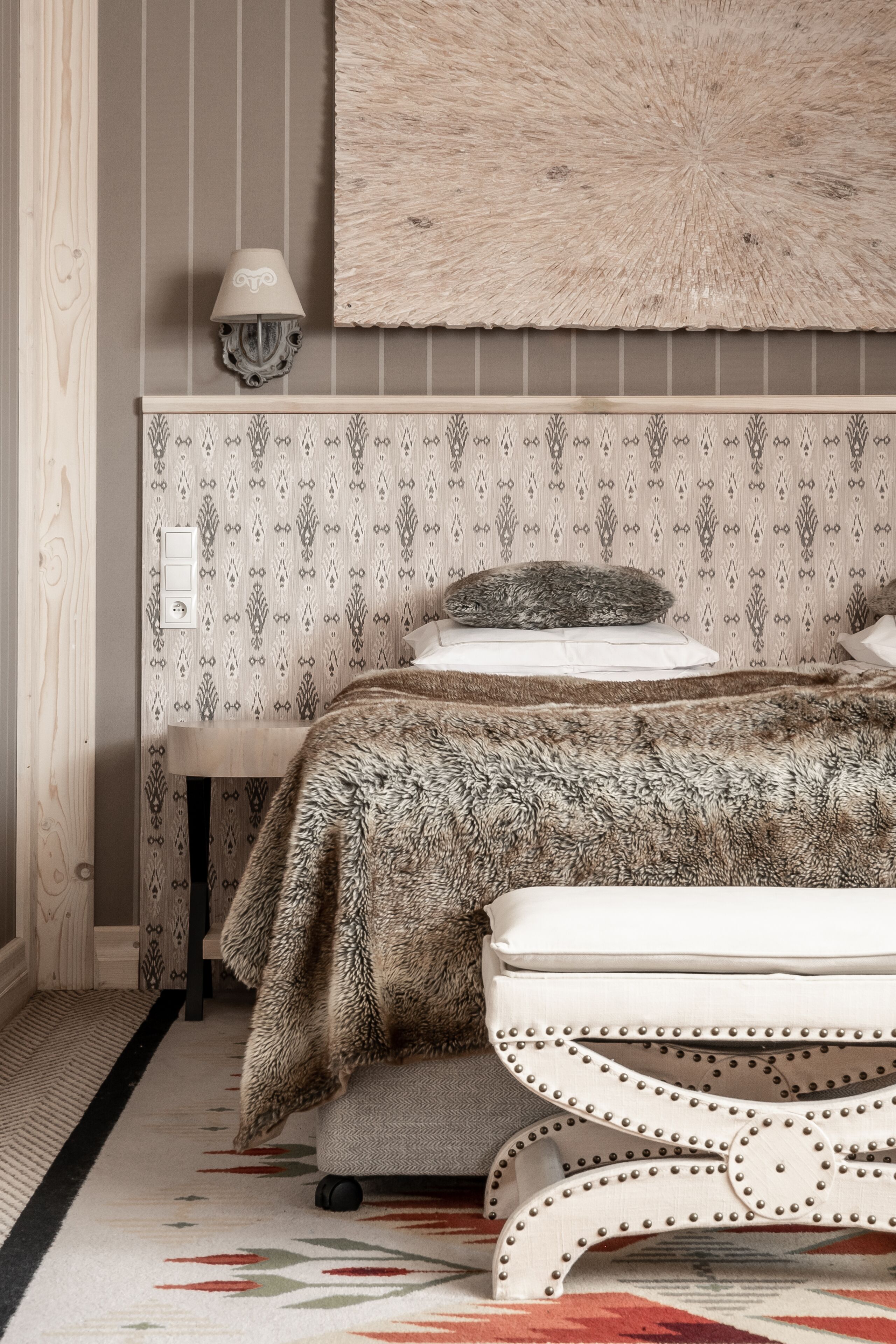 Quarto com cabeceira estampada, mesa de cabeceira, luminária, obra de arte abstrata grande, tapete estampado e um banco com design exclusivo.