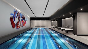 Piscina interior moderna com pistas bem definidas, cadeiras de descanso e uma grande obra de arte abstrata na parede.