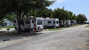 Vários trailers estacionados ao longo de uma estrada de cascalho, com árvores e uma casa ao fundo.
