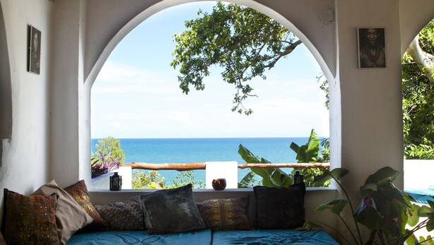 Quarto com vista para o mar através de uma janela em arco, cama com roupa de cama estampada e uma planta no parapeito.