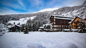 Estância de montanha coberta de neve com vários edifícios, incluindo uma grande hospedaria com varandas e uma cabana menor.