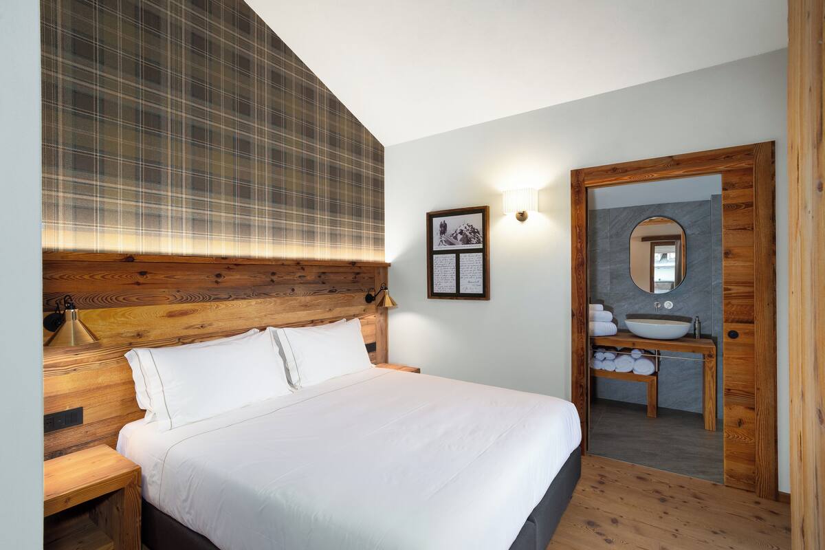 Quarto com cabeceira de madeira, cama com roupa de cama branca, luminária de cabeceira, quadro na parede e banheiro com toucador e pia de madeira.