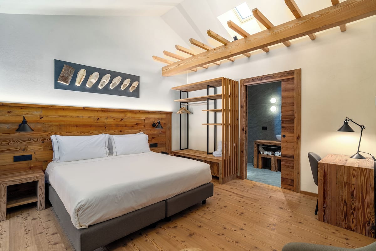 Quarto com cama, cabeceira de madeira, um banco de madeira, uma mesa pequena e um espelho.