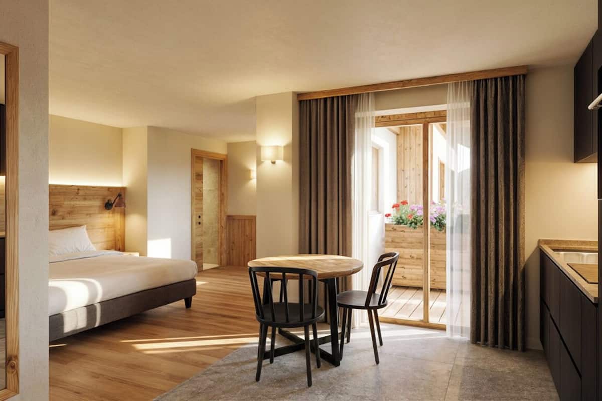 Quarto de hotel moderno com cama, mesa de jantar com cadeiras, cozinha e porta de vidro deslizante que dá para uma área externa.