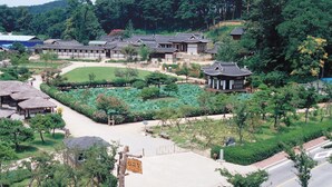 Um complexo de edifícios em estilo tradicional coreano, com várias construções, rodeado por vegetação exuberante e um jardim bem cuidado.